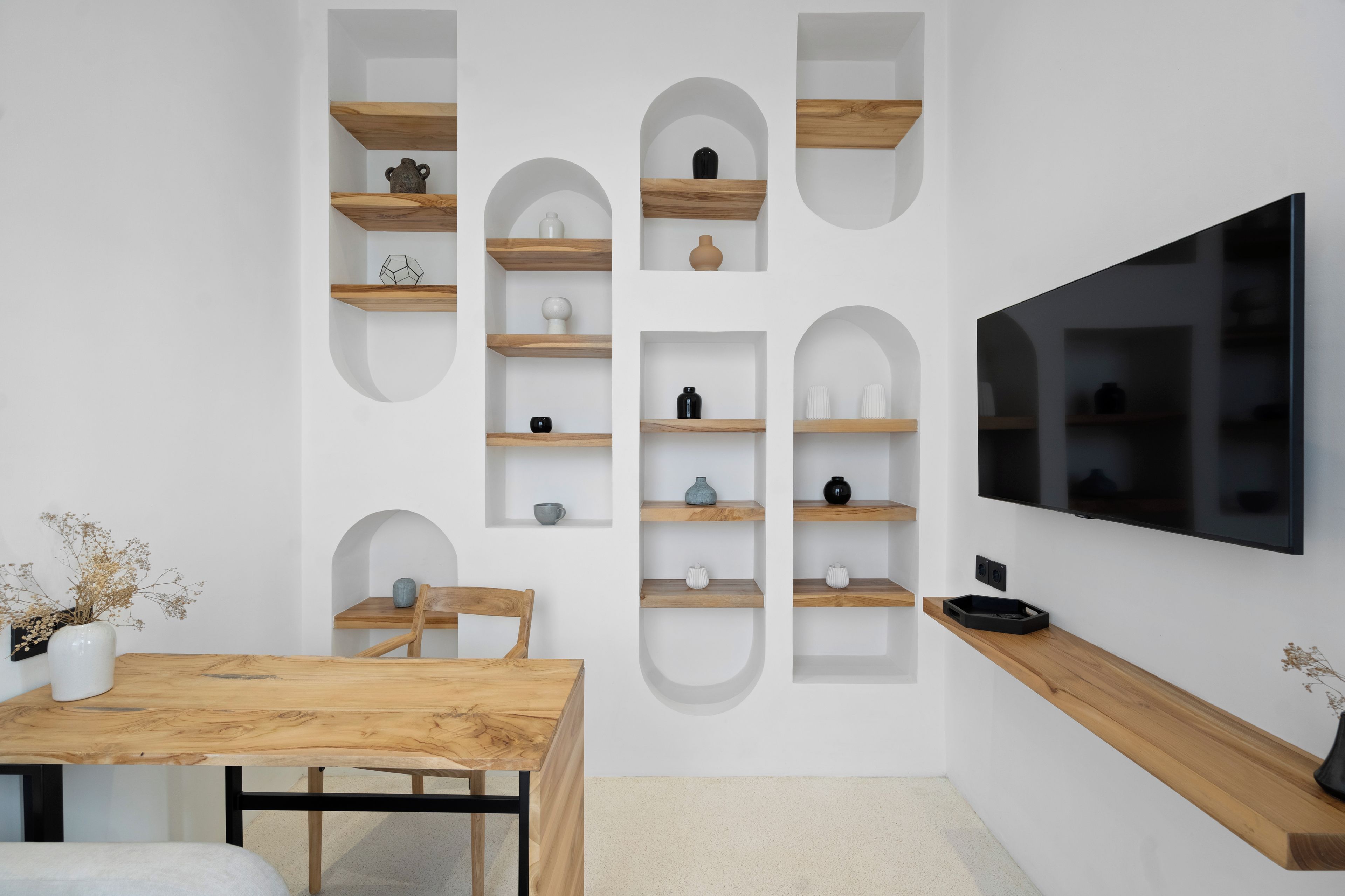 Uma sala de estar moderna com uma mesa de jantar de madeira, uma TV fixada na parede e estantes embutidas com diversos objetos decorativos.