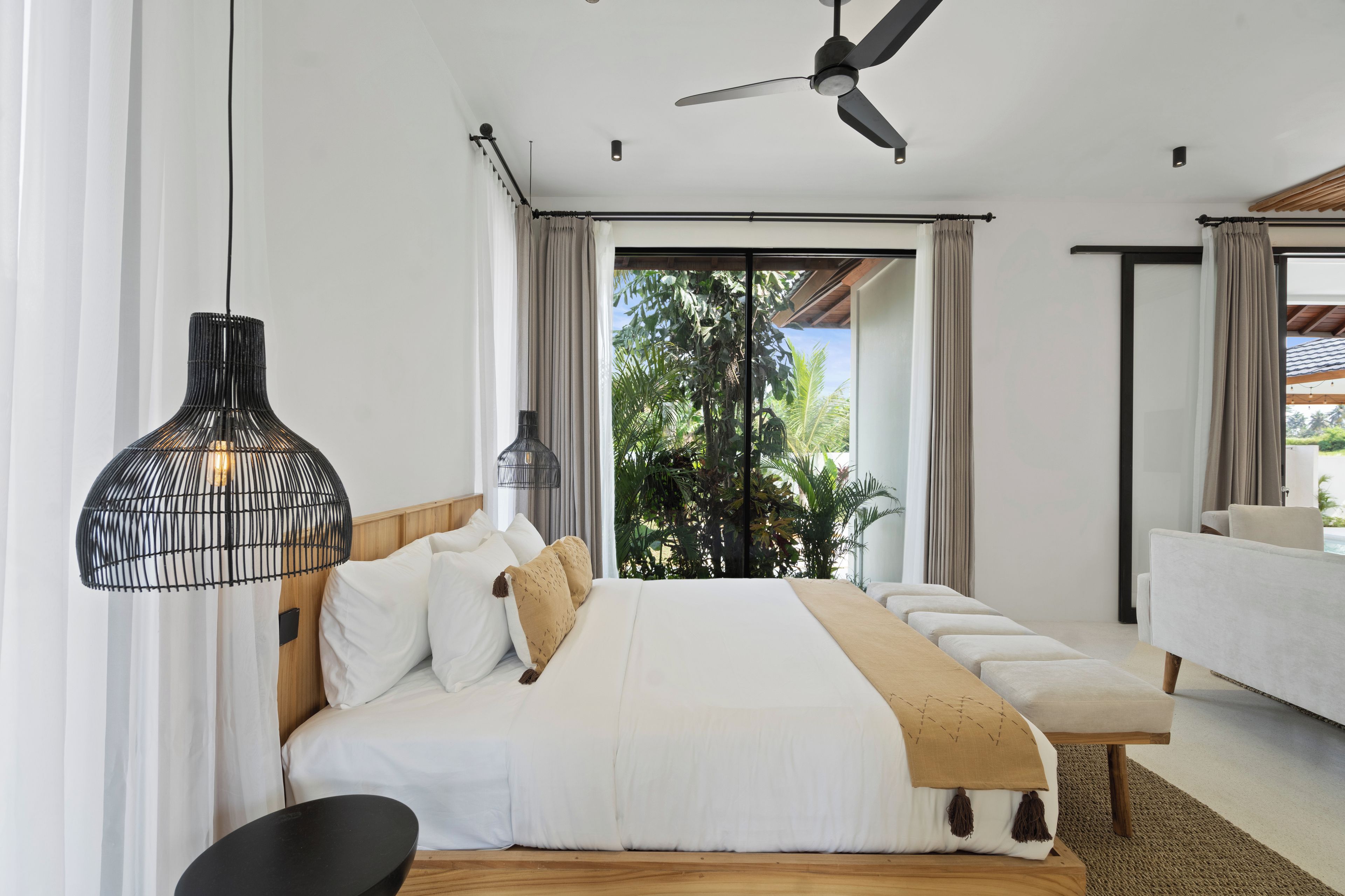 Quarto com cama, mesa de cabeceira, ventoinha no teto e vista para o verde lá fora.