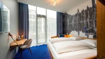Quarto de hotel com uma cama grande, uma escrivaninha com cadeira, televisão e um mural com cena de floresta.