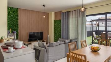 Uma sala de estar moderna com televisão de tela plana, sofá cinza, mesa de jantar com cadeiras e uma varanda com espaço para sentar ao ar livre.