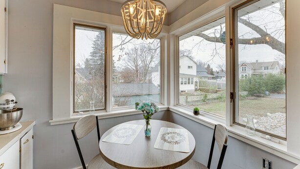Uma sala de jantar com uma mesa redonda, duas cadeiras, um candelabro e janelas amplas com vista para o bairro.