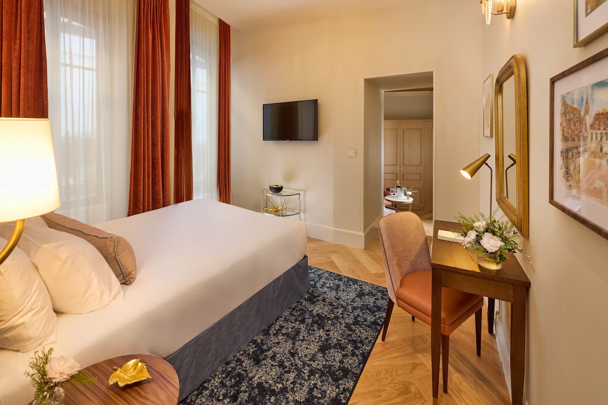 Quarto de hotel com cama, escrivaninha, cadeira, televisão, espelho e janela com cortinas.