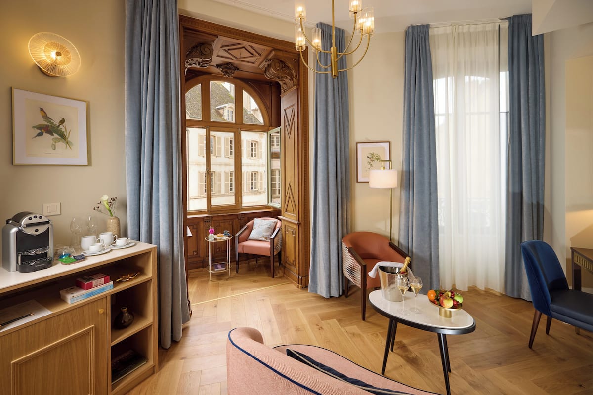 Quarto de hotel acolhedor com uma secretária de madeira, uma poltrona confortável, uma pequena mesa com um vaso e frutas, e vista para um edifício através de uma janela grande.