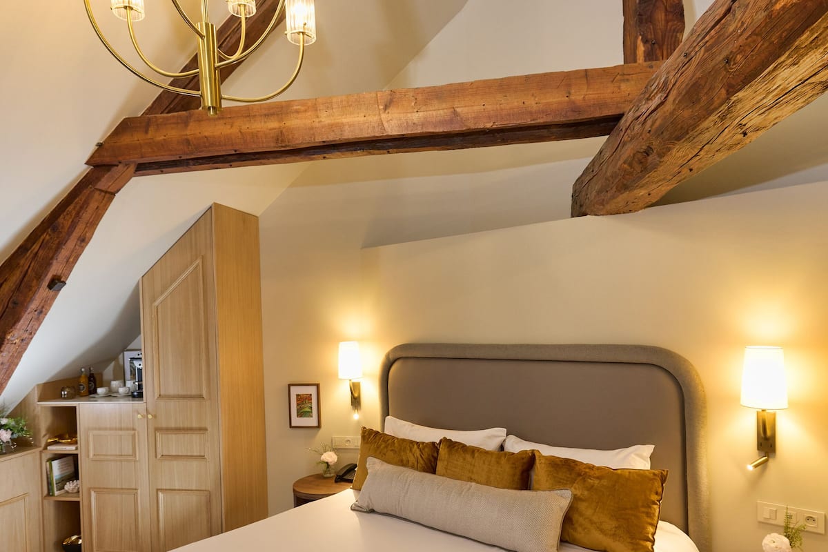 Quarto com cama grande, luminária, vigas de madeira, mesa de cabeceira com um vaso e um pequeno armário.