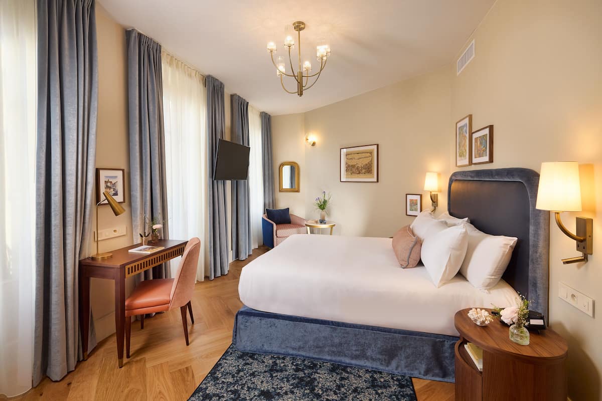 Quarto de hotel com uma cama grande, uma escrivaninha com cadeira, uma televisão e um candelabro.