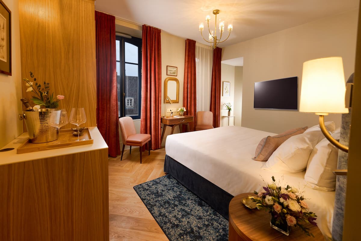 Quarto de hotel com uma cama grande, uma escrivaninha com luminária, uma cadeira, uma pequena mesa com um vaso de flores e uma TV fixada na parede.
