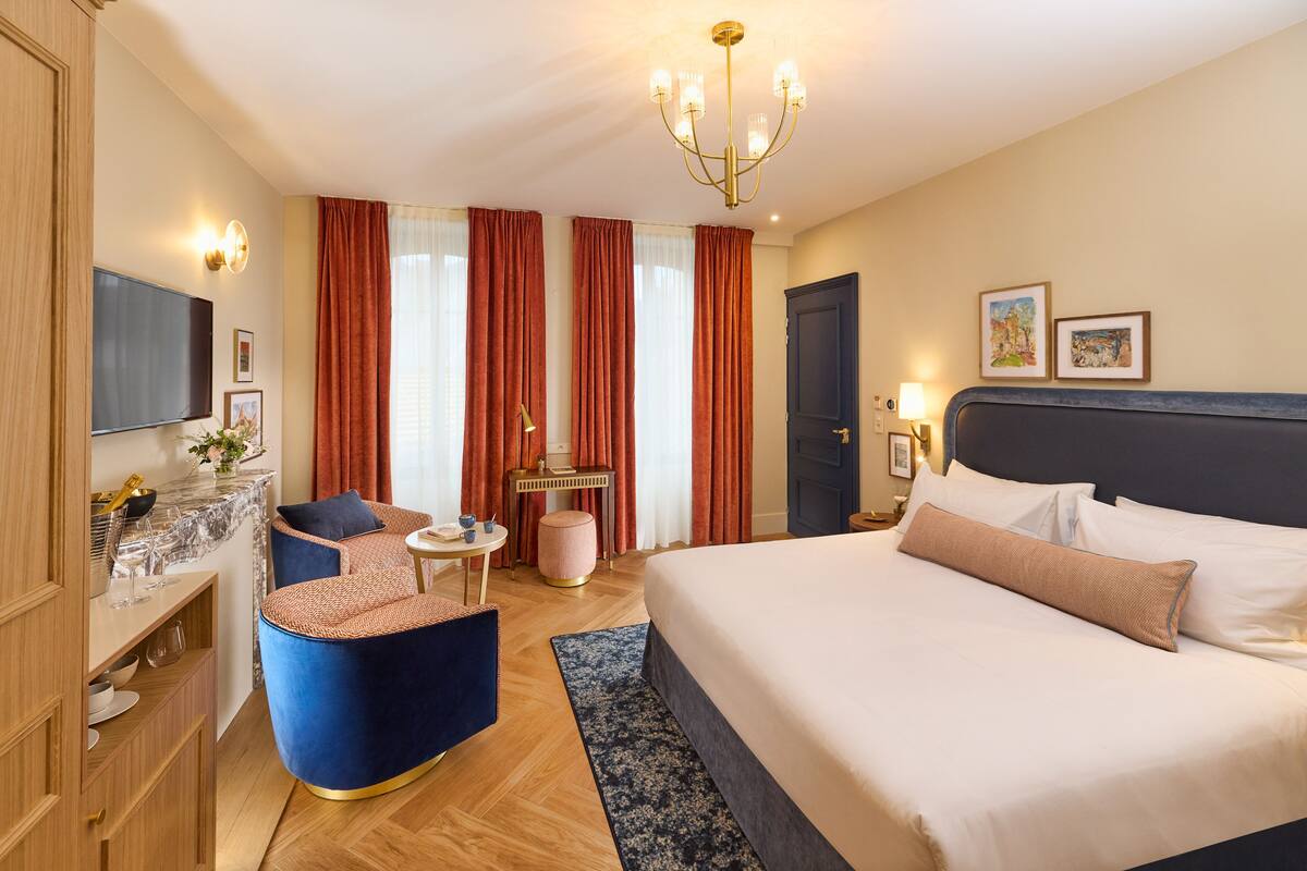 Quarto de hotel com uma cama grande, uma poltrona azul, uma pequena mesa, uma televisão e um candelabro.