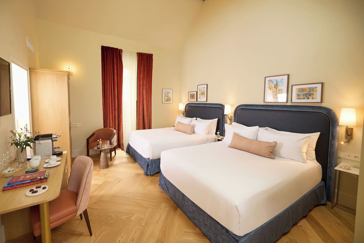 Quarto de hotel com duas camas, uma escrivaninha com cadeira, uma mesinha e uma janela com cortinas.