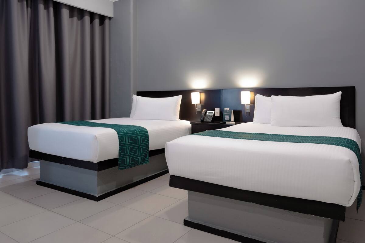 Quarto de hotel com duas camas, cada uma com lençóis brancos e manta verde estampada. Há duas mesas de cabeceira com luminárias, um telefone e uma pequena mesa com bandeja. O quarto possui cortina cinza e piso de azulejo branco.