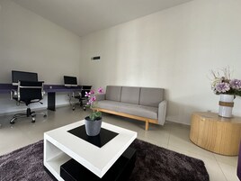 Uma sala de estar moderna com um sofá cinza, uma cadeira de escritório preta, uma mesa de centro branca e uma pequena mesa de apoio de madeira com um vaso de flores.