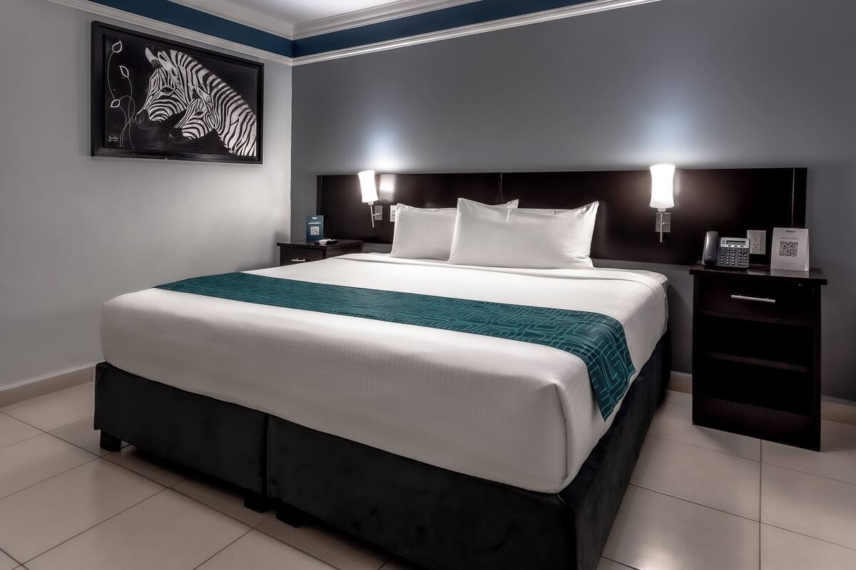 Quarto de hotel moderno com uma cama grande, cabeceira com iluminação embutida, mesa de cabeceira com telefone e um código QR, e uma obra de arte emoldurada na parede.