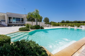 Uma área de piscina moderna com água azul cristalina, rodeada por vegetação exuberante e um edifício contemporâneo com grandes janelas.