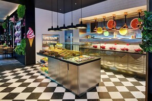 Uma gelataria moderna com chão quadriculado, um balcão central de atendimento e uma área de cozinha decorada com frutas cítricas penduradas.