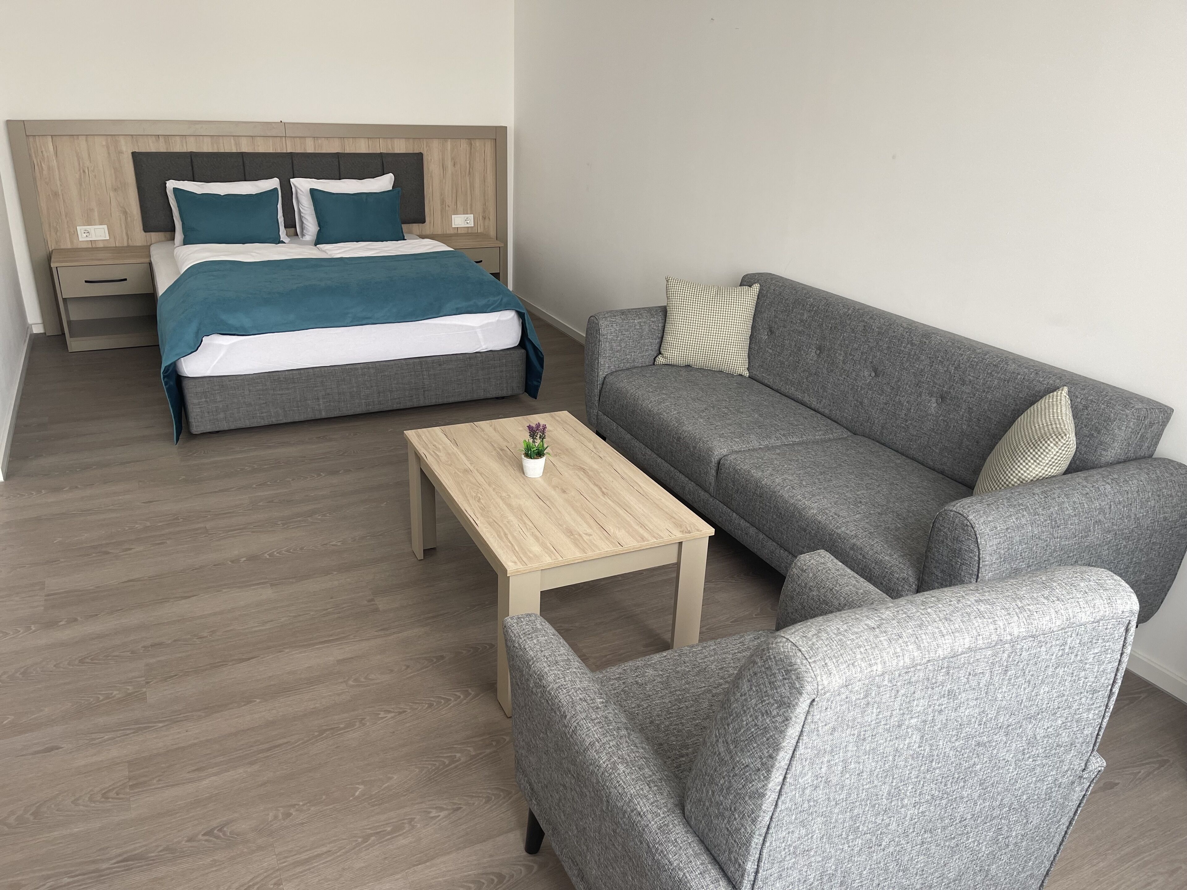Quarto com cama, sofá cinza, mesa pequena e mesa de cabeceira.