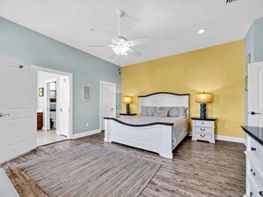 Quarto com cama, mesas de cabeceira, ventoinha no teto e piso de madeira.
