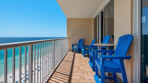 Uma varanda com cadeiras azuis e uma mesa, com vista para uma praia com guarda-sóis e pessoas.