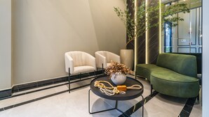 Um hall moderno com um sofá verde, duas poltronas brancas, uma mesa de centro redonda com um vaso e um colar de contas, e uma planta alta.