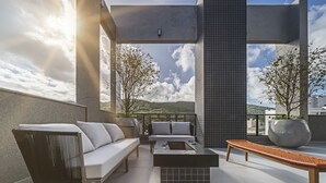 Uma área externa moderna com assentos, incluindo um sofá, um banco e uma mesa, cercada por janelas amplas e plantas em vasos.
