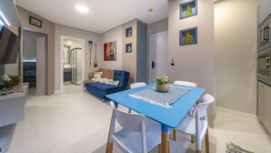 Sala de estar moderna com uma mesa azul, cadeiras brancas, um sofá azul e uma área de cozinha com geladeira e forno.