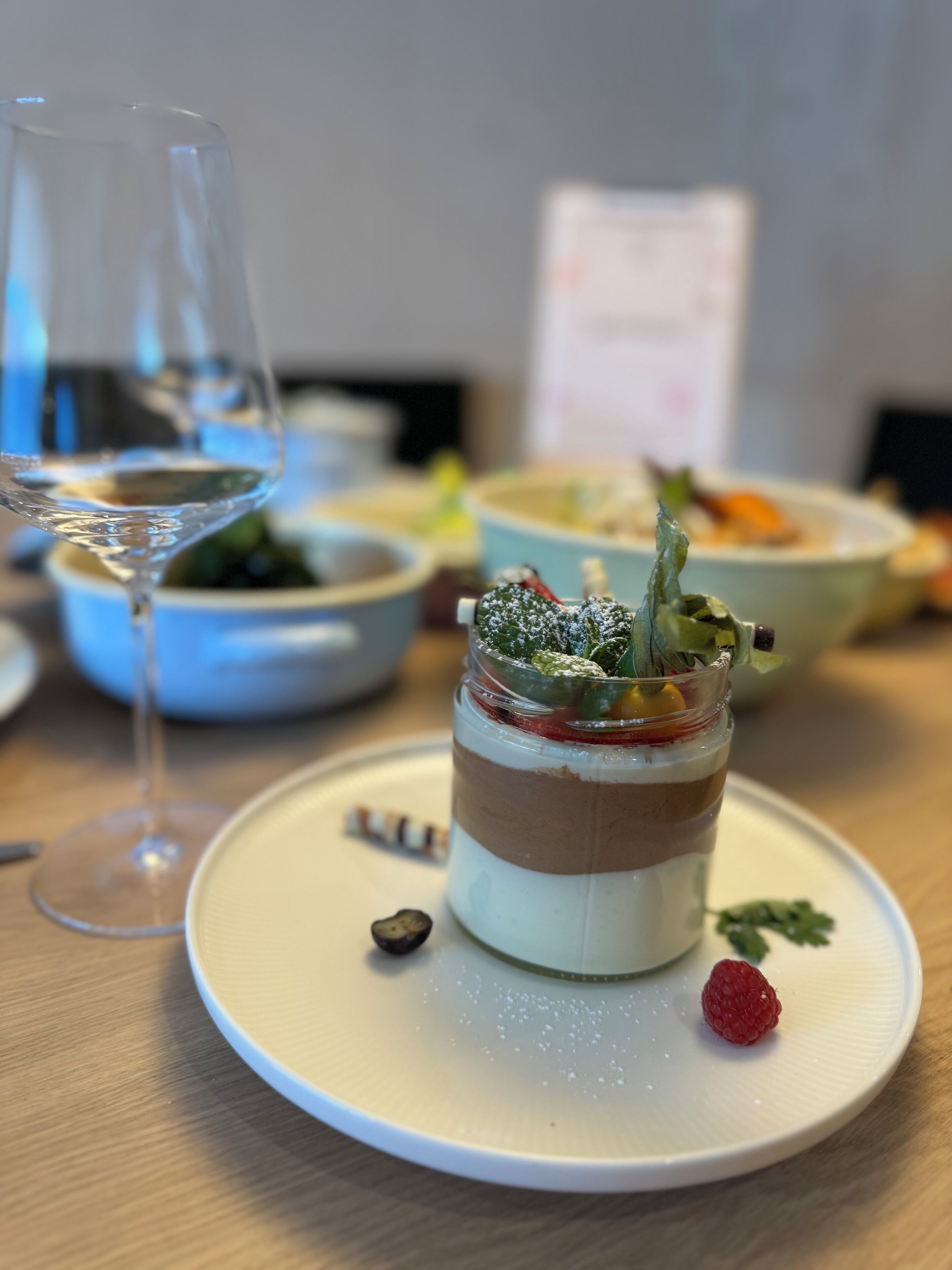 Sobremesa dentro de um pote de vidro sobre um prato, com um copo de água e uma saladeira ao fundo.