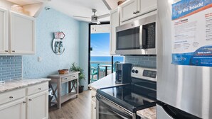Uma cozinha moderna com armários brancos, eletrodomésticos de aço inoxidável e vista para o oceano através de uma porta aberta.