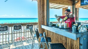 Um bar à beira-mar com vista para o oceano, cadeiras de metal e um bartender preparando bebidas.