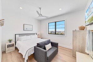 Um quarto com cama, poltrona cinza, mesa de cabeceira com planta, ventoinha no teto, janela com vista e guarda-roupa de madeira.