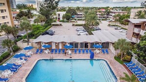 Piscina com espreguiçadeiras e guarda-sóis, cercada por um estacionamento e edifícios.