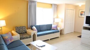 Sala de estar com sofá, poltrona e uma mesinha. Há uma janela com cortinas, uma televisão e um armário.