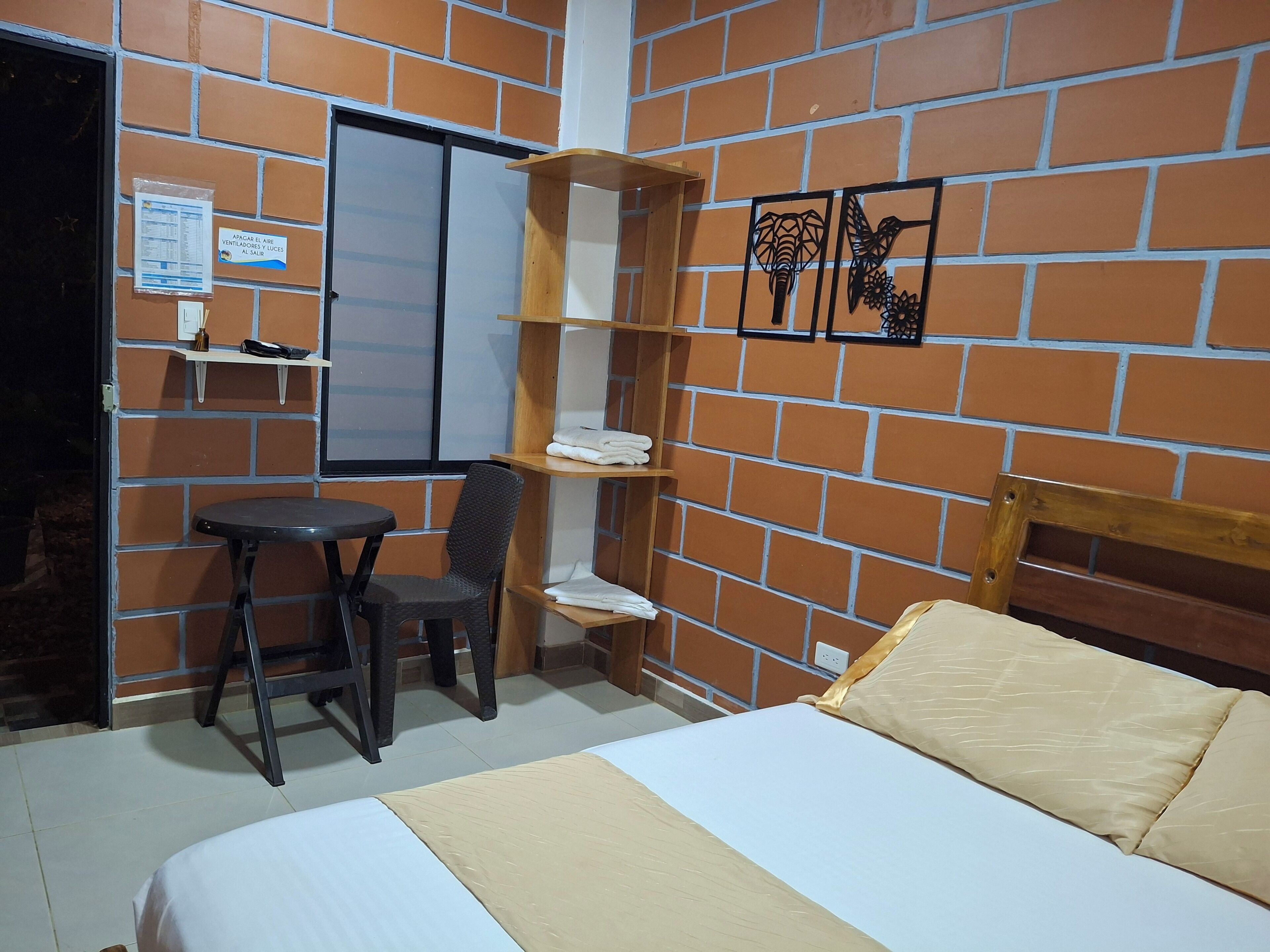 Quarto com uma cama, uma pequena mesa, uma cadeira e uma estante com toalhas.
