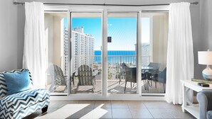 Quarto de hotel moderno com uma grande porta de vidro deslizante que dá para uma varanda com mobiliário exterior e vista para o oceano e edifícios.