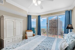 Quarto com cama, guarda-roupa, cadeira e varanda com vista para o oceano e edifícios.