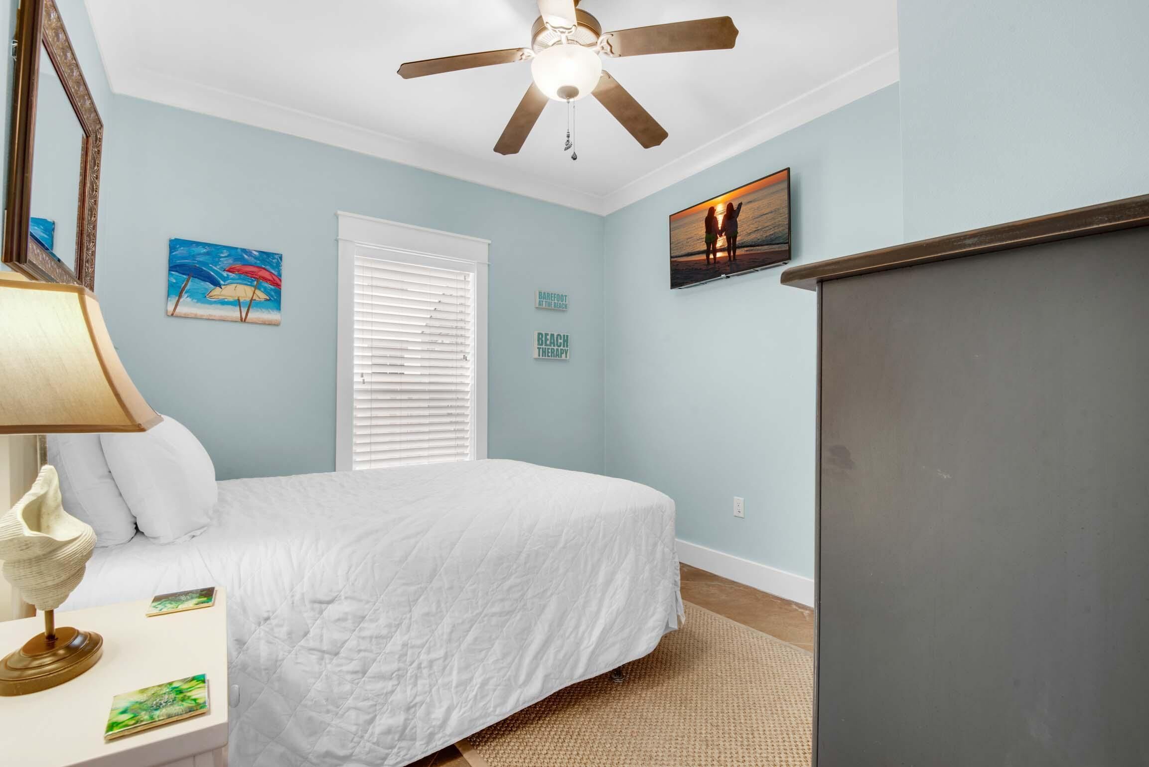 Quarto com cama, ventilador de teto, escrivaninha com luminária e obras de arte nas paredes.