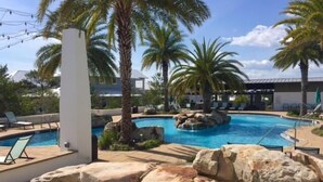 Área de piscina com palmeiras, espreguiçadeiras e um edifício ao fundo.