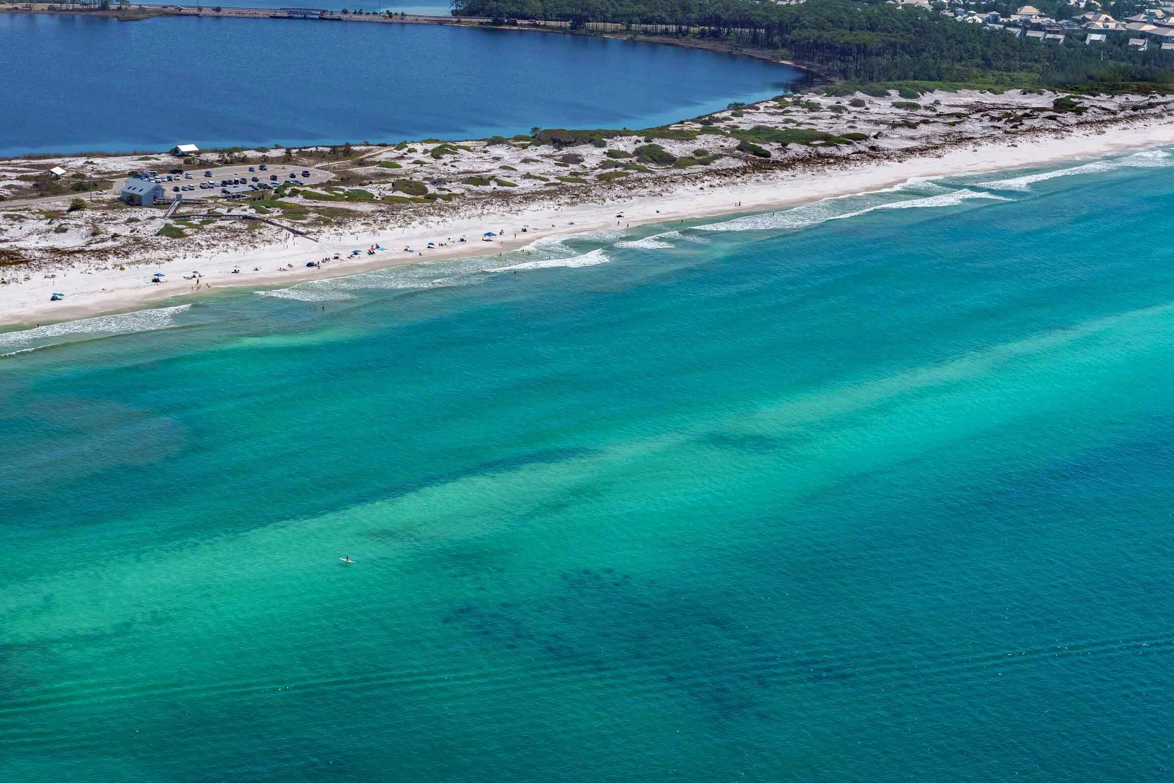 Vista aérea de uma praia com águas turquesa cristalinas, faixa de areia e um pequeno edifício com telhado azul.