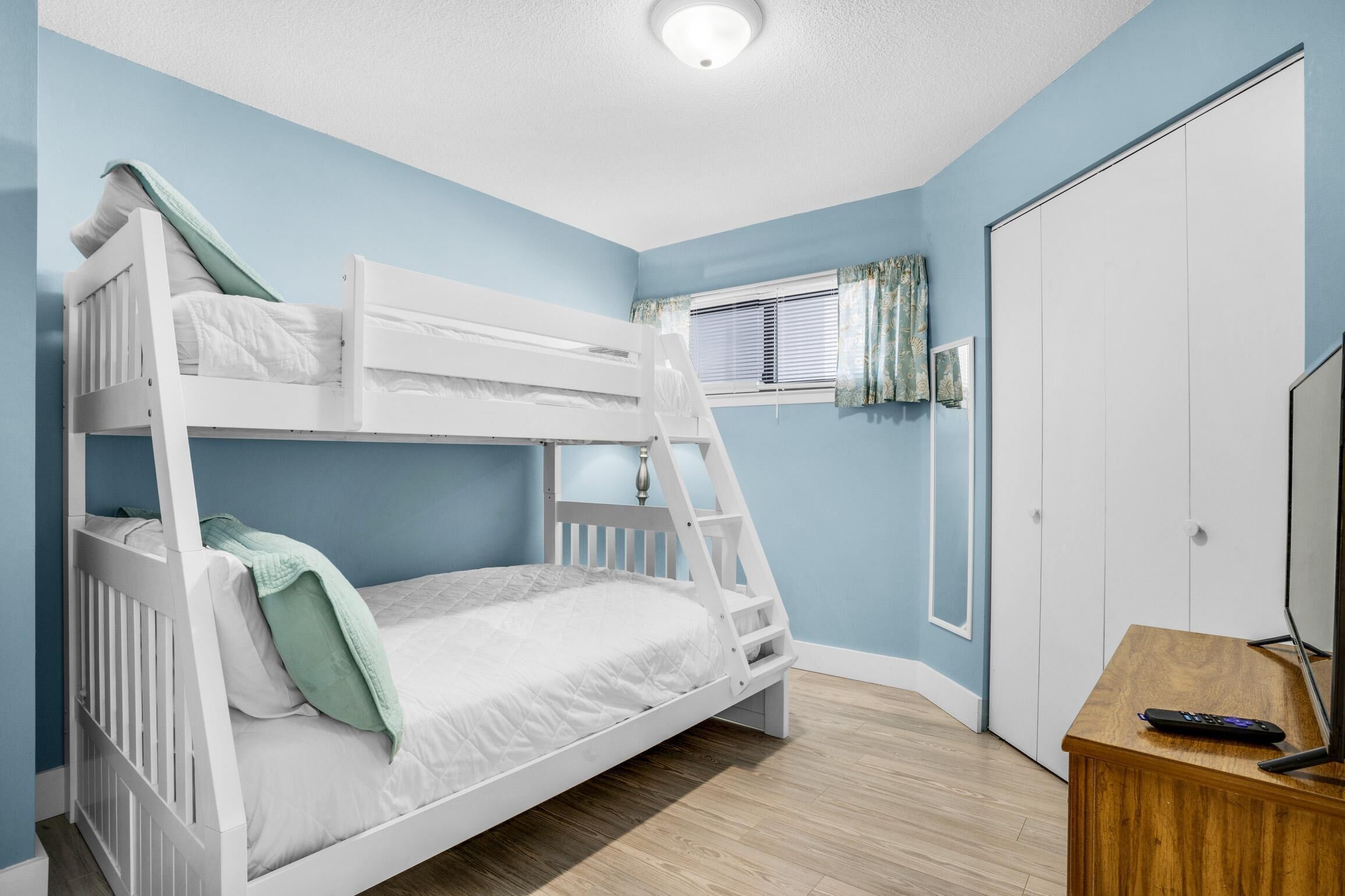 Uma cama beliche com roupa de cama branca e um cobertor verde, uma escrivaninha de madeira com um controle remoto e um guarda-roupa branco em um quarto com paredes azuis e piso de madeira.
