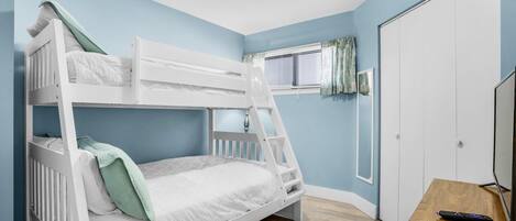 Uma cama beliche com roupa de cama branca e um cobertor verde, uma escrivaninha de madeira com um controle remoto e um guarda-roupa branco em um quarto com paredes azuis e piso de madeira.