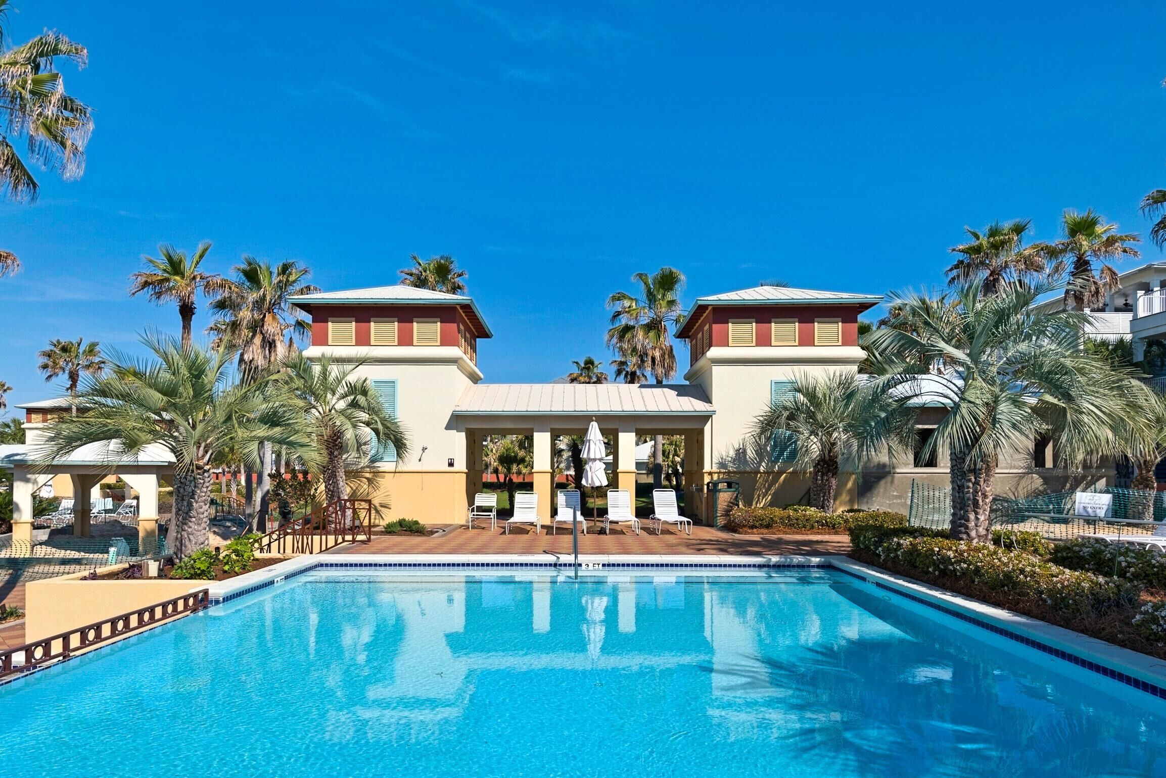 Piscina com espreguiçadeiras e palmeiras, cercada por edifícios com arquitetura mediterrânea.
