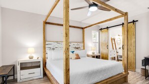 Quarto com cama de dossel, vigas de madeira, ventilador de teto, mesa de cabeceira com abajur, uma pequena mesa e uma cadeira.