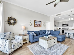 Uma sala de estar com um sofá azul, uma poltrona estampada, uma mesa de centro de madeira, um abajur e uma guirlanda decorativa na parede.