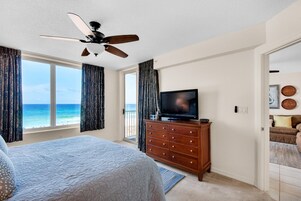Quarto com cama, televisão sobre uma cômoda de madeira, ventilador de teto e vista para o oceano através de grandes janelas.