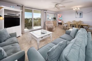 Sala de estar com sofá cinzento, mesa de centro branca, televisão em um móvel branco, área de jantar com mesa e cadeiras, e varanda com vista.