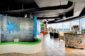 Área interna de piscina moderna com parede em mosaico, um letreiro com a palavra 'SPLASH' e vista para o oceano através de amplas janelas.
