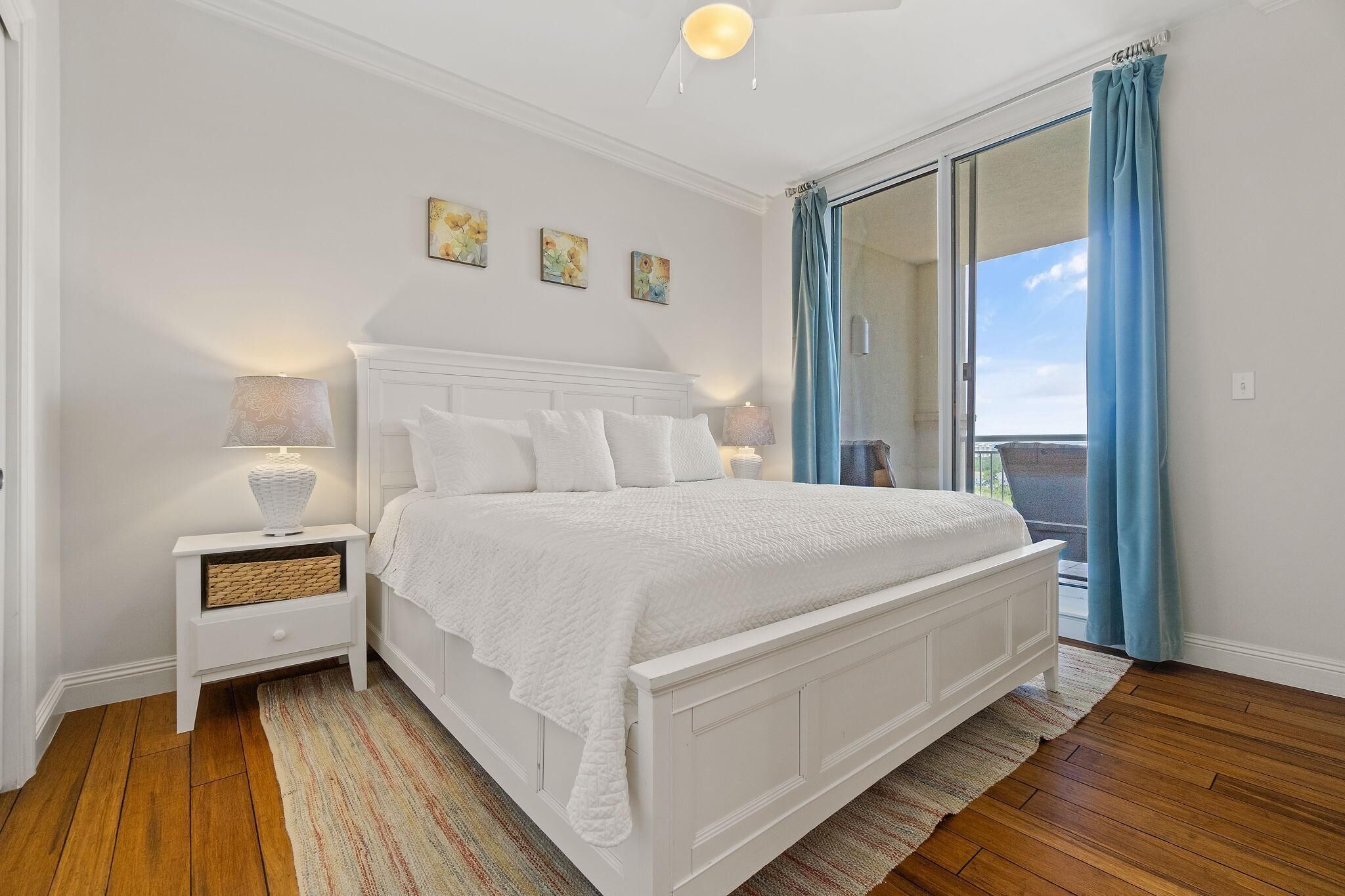 Quarto com cama branca, mesa de cabeceira com abajur e vista para o exterior através de uma porta deslizante.