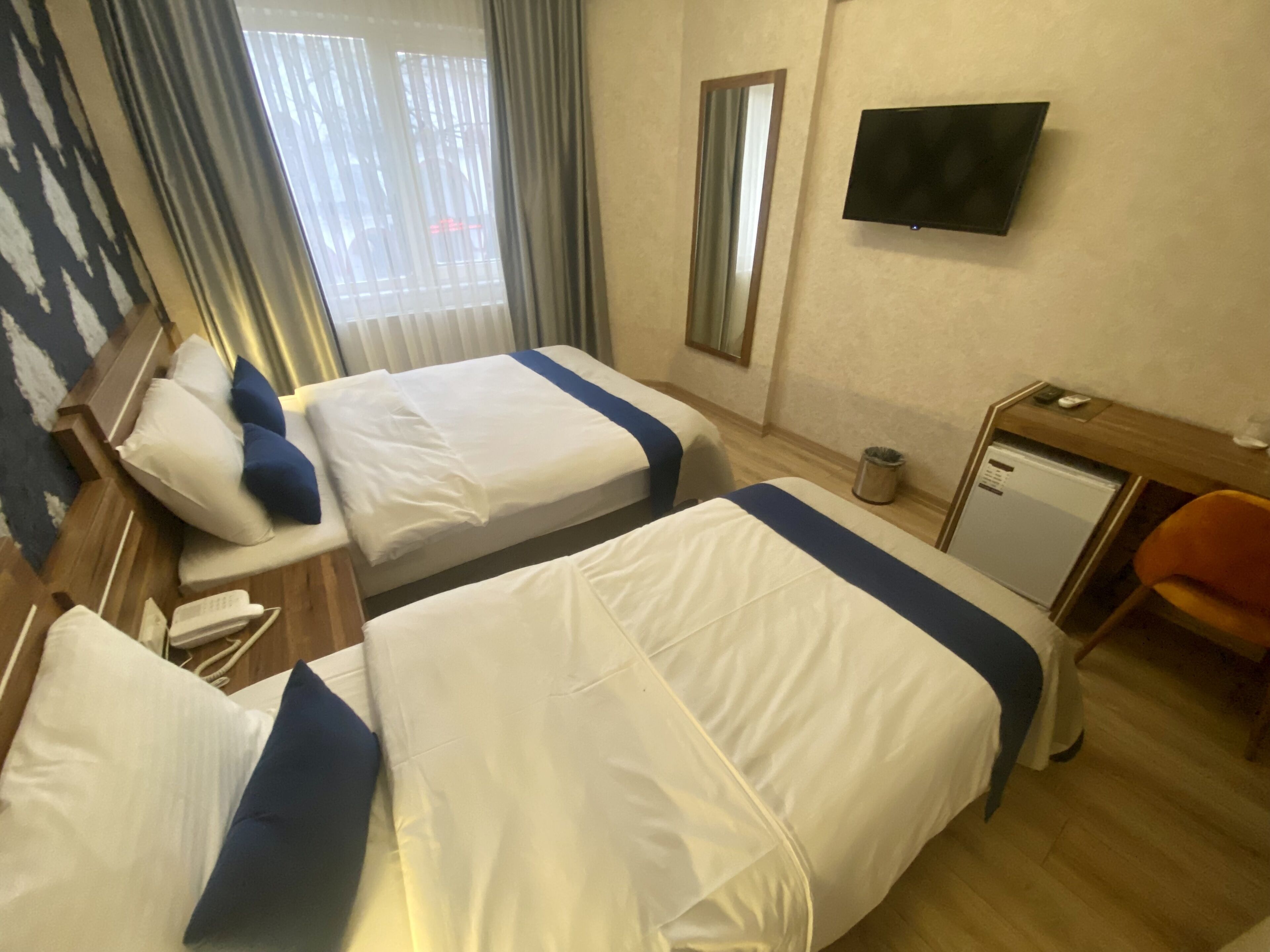 Quarto de hotel com duas camas, uma mesa de escritório com computador, uma cadeira, televisão e janela com cortinas.
