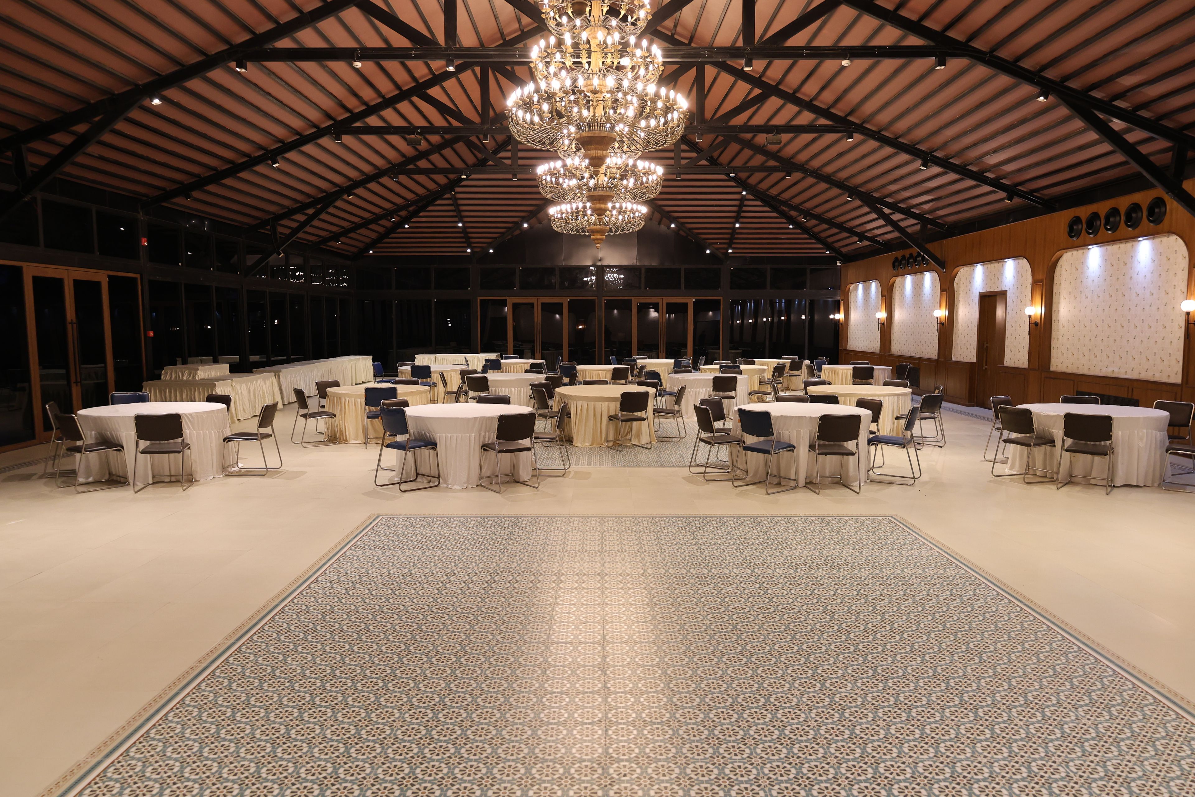 Um amplo salão de banquetes com várias mesas redondas e cadeiras, um tapete estampado e um grande candelabro.