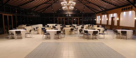Um amplo salão de banquetes com várias mesas redondas e cadeiras, um tapete estampado e um grande candelabro.