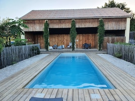 Um deck de madeira com uma piscina retangular, cercado por um muro de madeira e um edifício de madeira com telhado em telhas.
