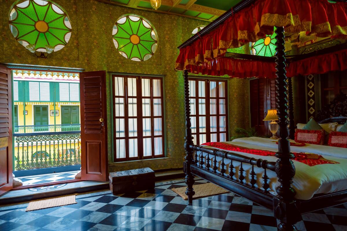 Uma cama de dossel com dossel vermelho, chão quadriculado e janelas grandes com vidros verdes.