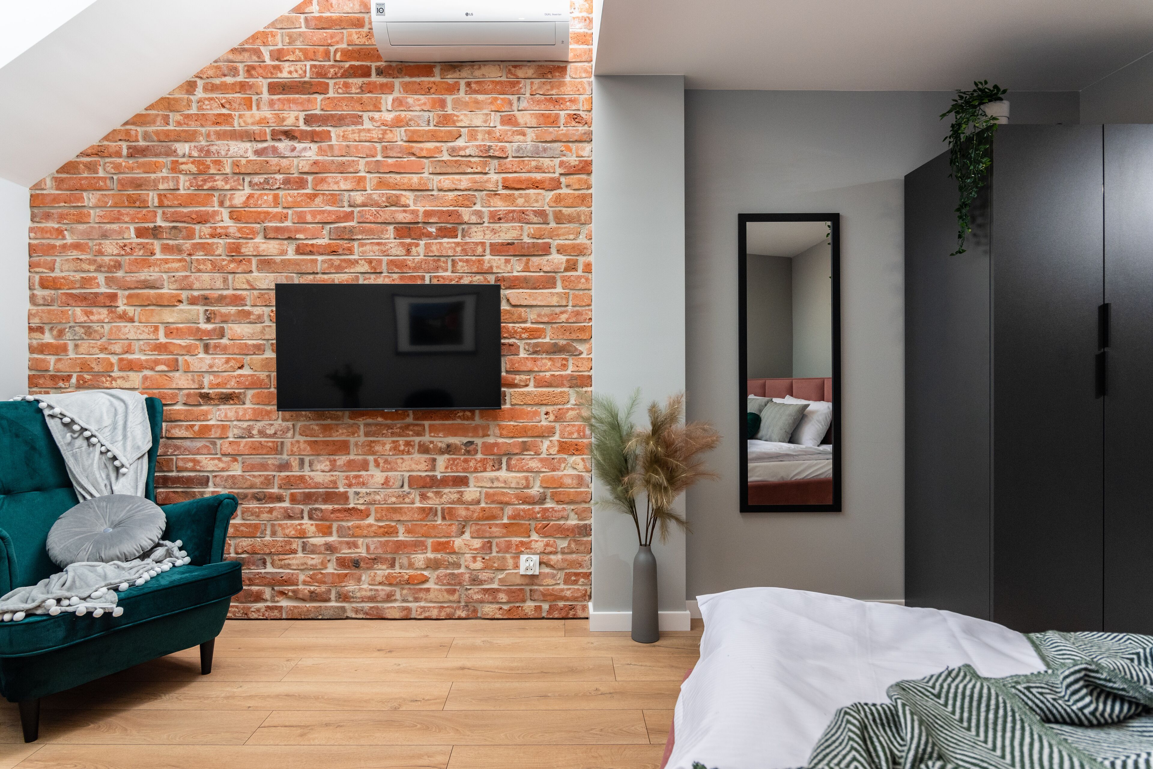 Quarto moderno com parede de tijolos, TV de tela plana, cama, espelho e poltrona verde.